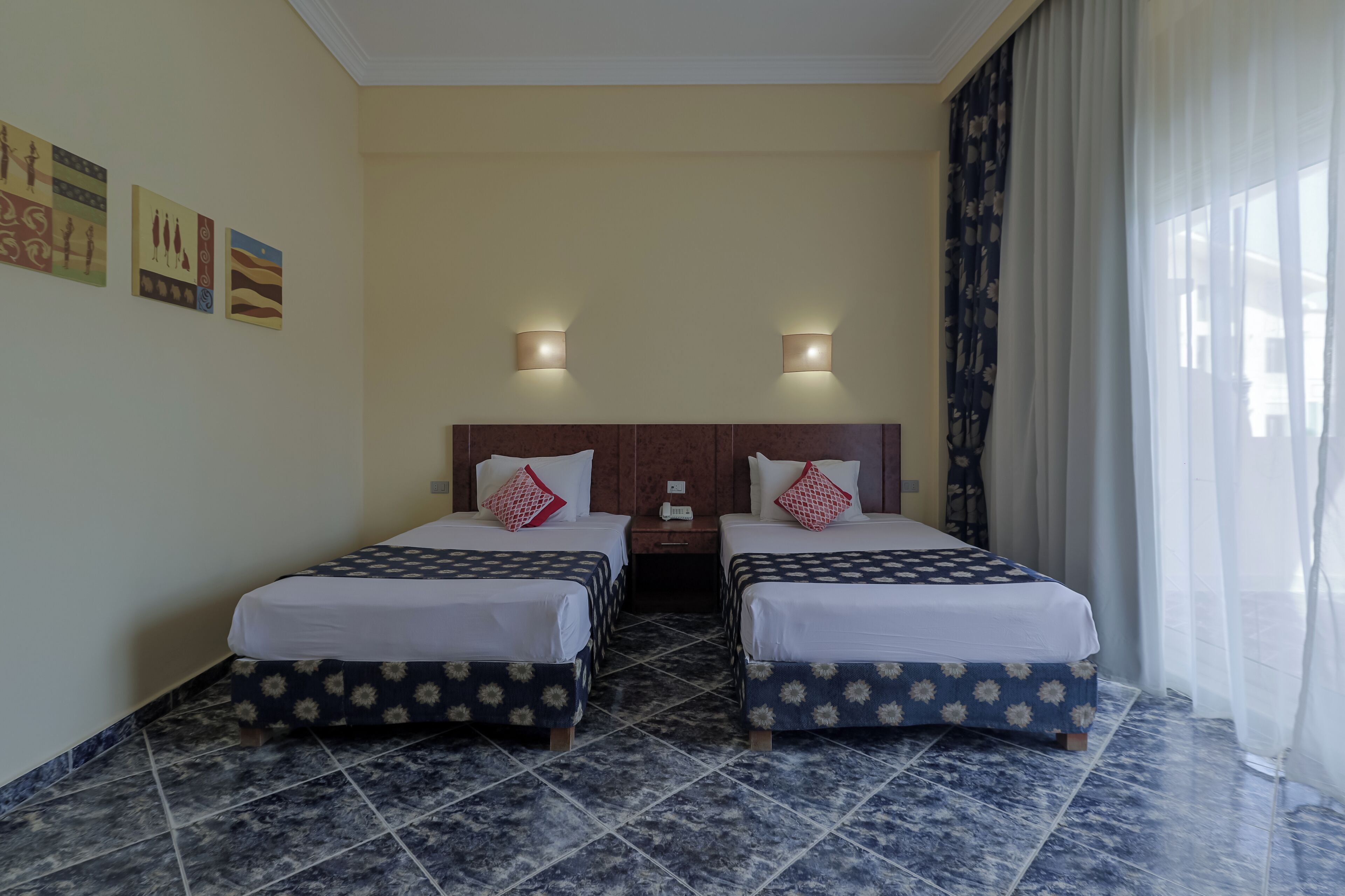 Quarto de hotel com duas camas, cabeceira, luminárias fixadas na parede e janela com cortinas.
