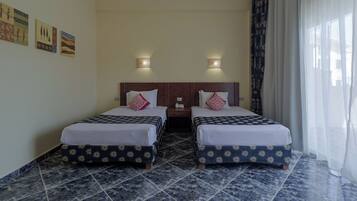 Quarto de hotel com duas camas, cabeceira, luminárias fixadas na parede e janela com cortinas.
