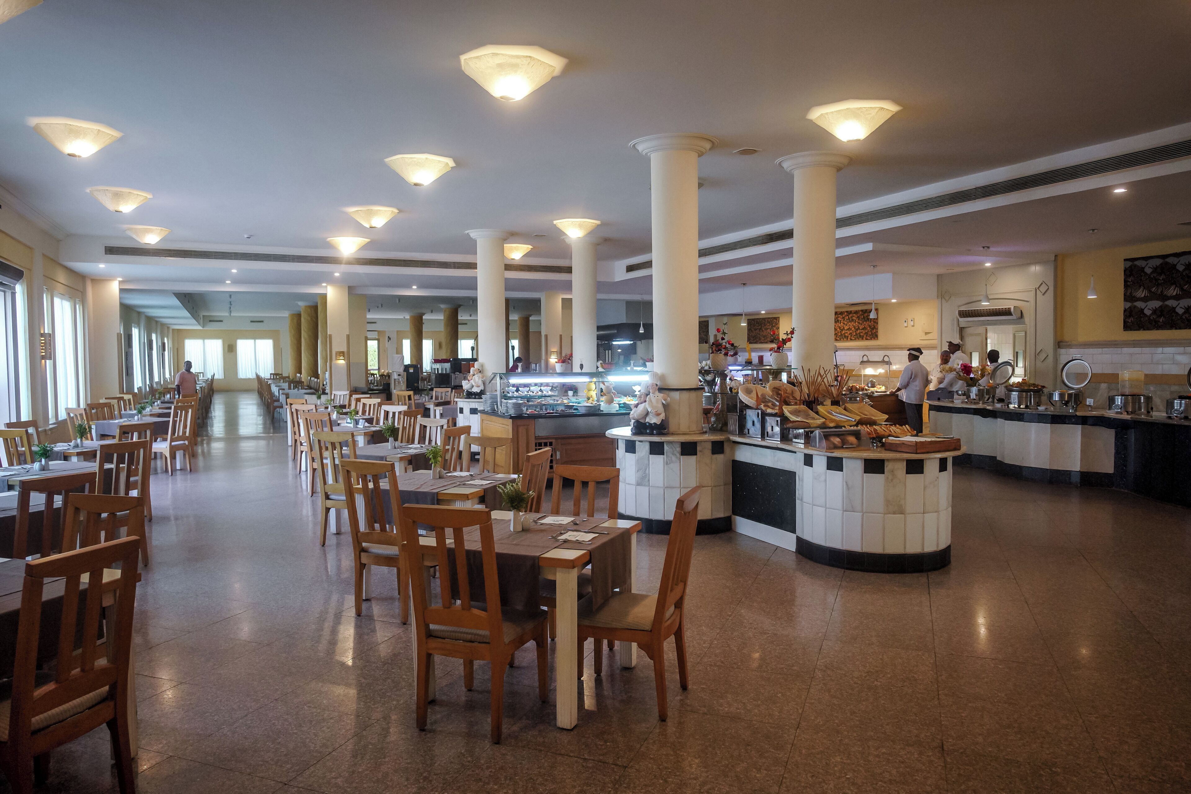 Ampla sala de jantar de hotel, com diversas mesas e cadeiras, um buffet e um longo corredor com janelas.