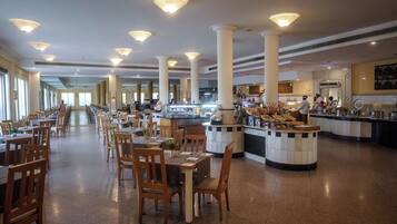 Ampla sala de jantar de hotel, com diversas mesas e cadeiras, um buffet e um longo corredor com janelas.