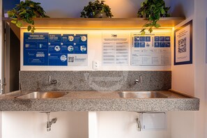 Casa de banho pública com bancadas de granito, duas pias e cartazes informativos na parede.
