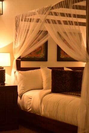 Uma cama com roupa de cama branca e um dossel, uma mesinha de cabeceira com abajur, e duas fotos emolduradas na parede.