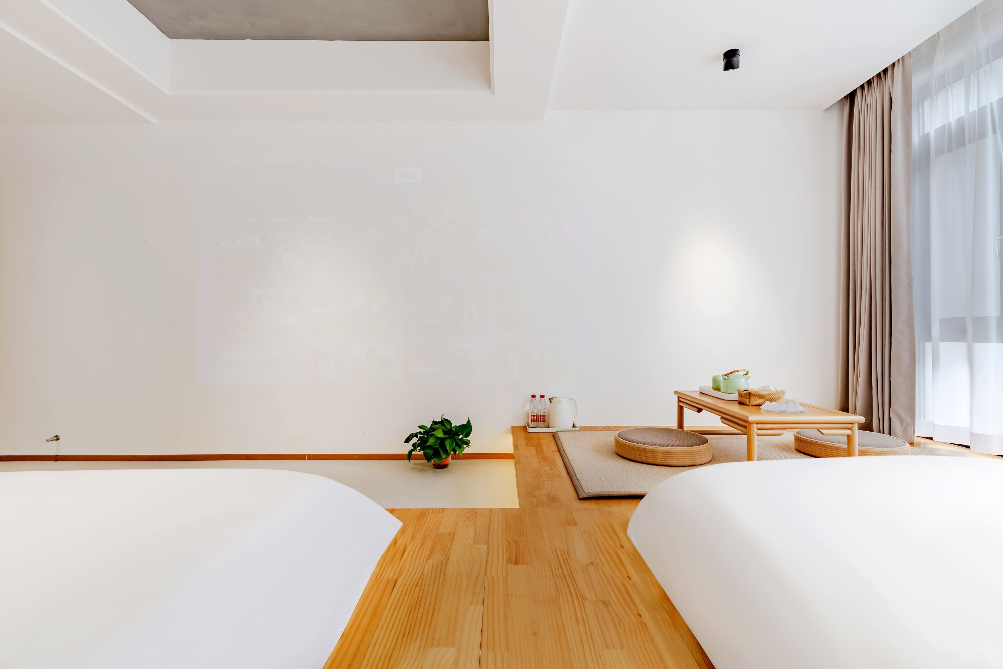 Quarto de hotel moderno com chão de madeira, duas camas, uma mesinha com uma planta e uma janela com cortinas.