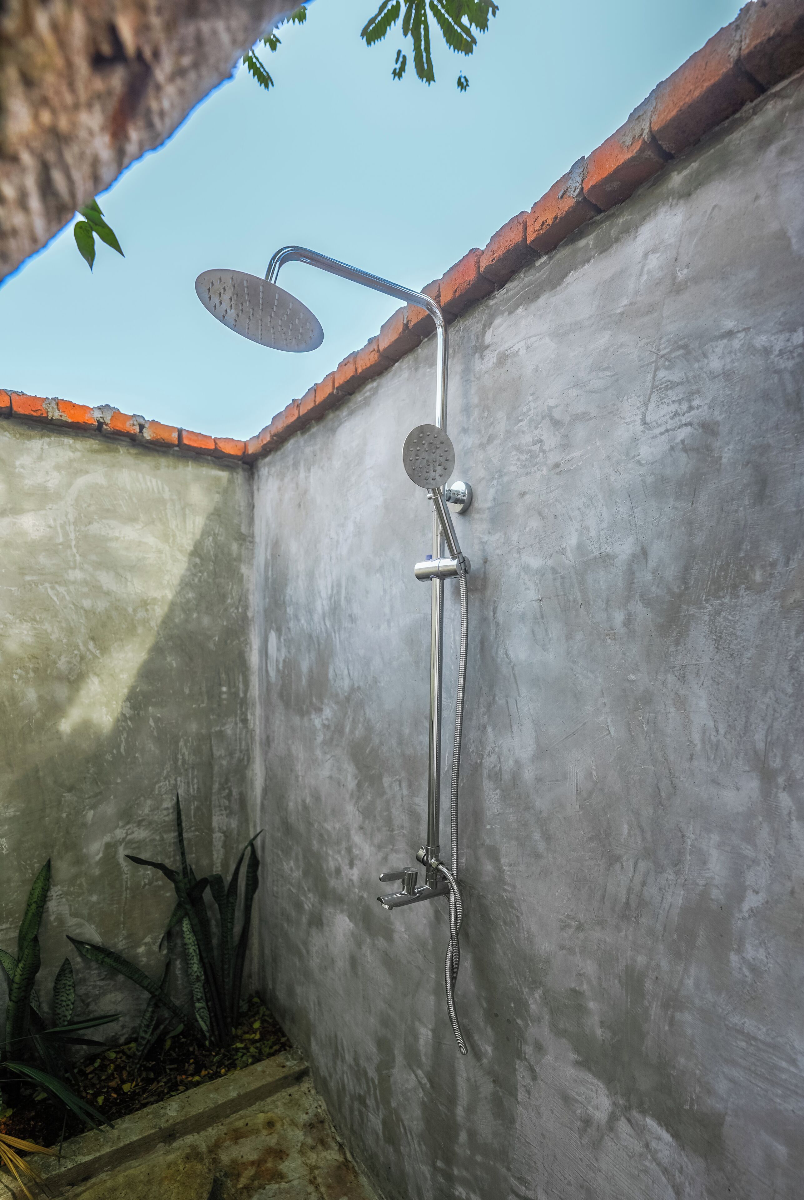 Compartimento de chuveiro em concreto com chuveiro e mangueira de metal, um nicho de tijolos e plantas verdes.