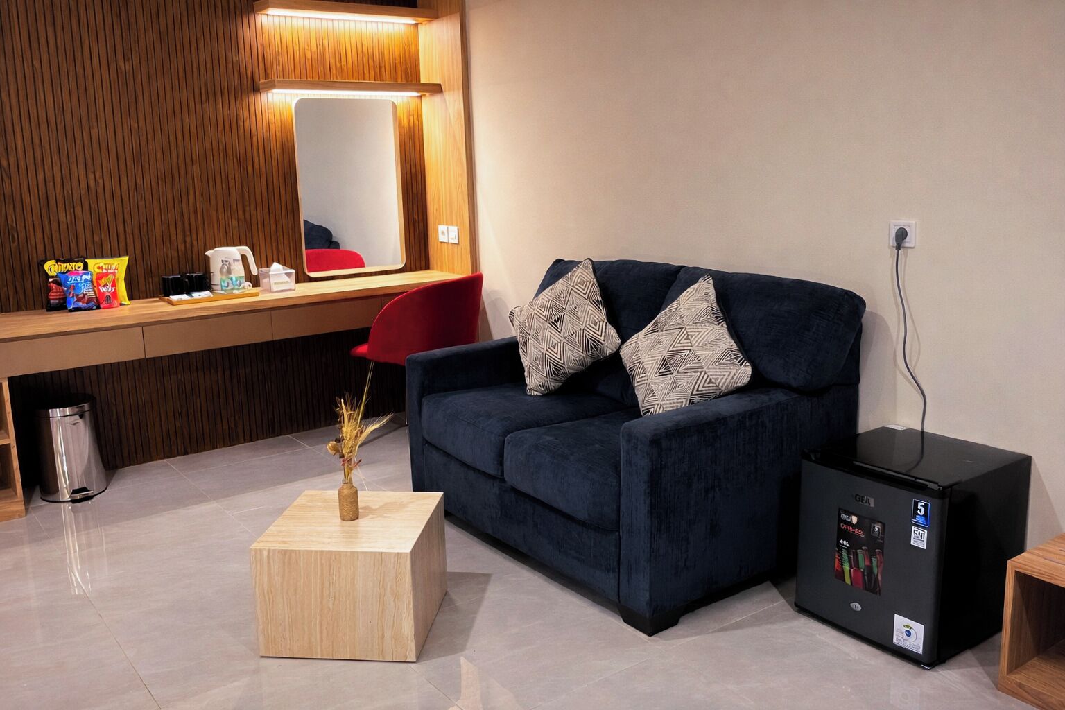 Uma sala de estar moderna com um sofá azul escuro, duas almofadas estampadas, uma mesa de centro de madeira com um vaso, uma escrivaninha com um espelho e um dispositivo eletrônico preto.