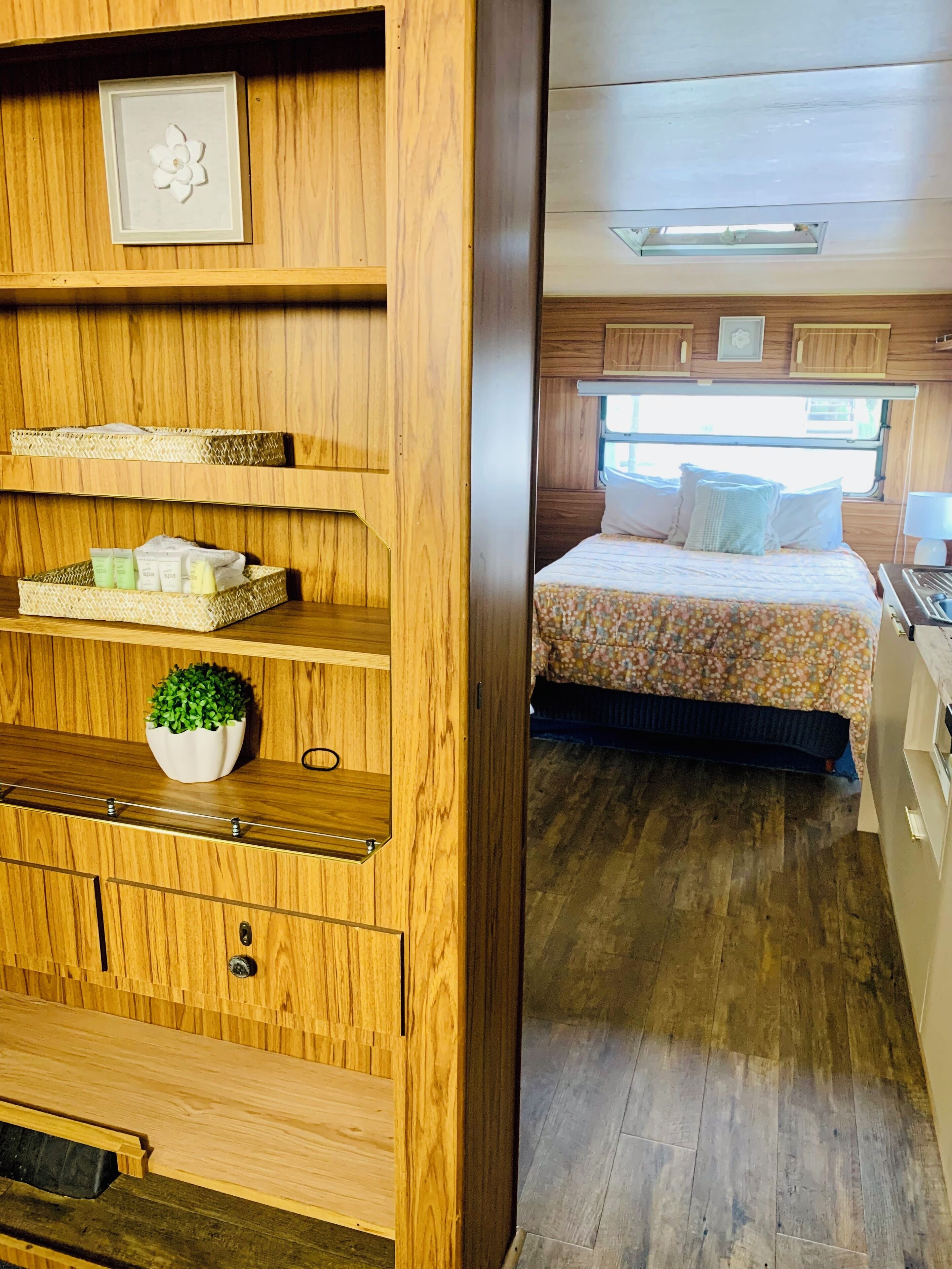 Interior compacto de um veículo recreativo, com decoração em madeira, uma cama com roupa de cama florida e uma pequena cozinha.