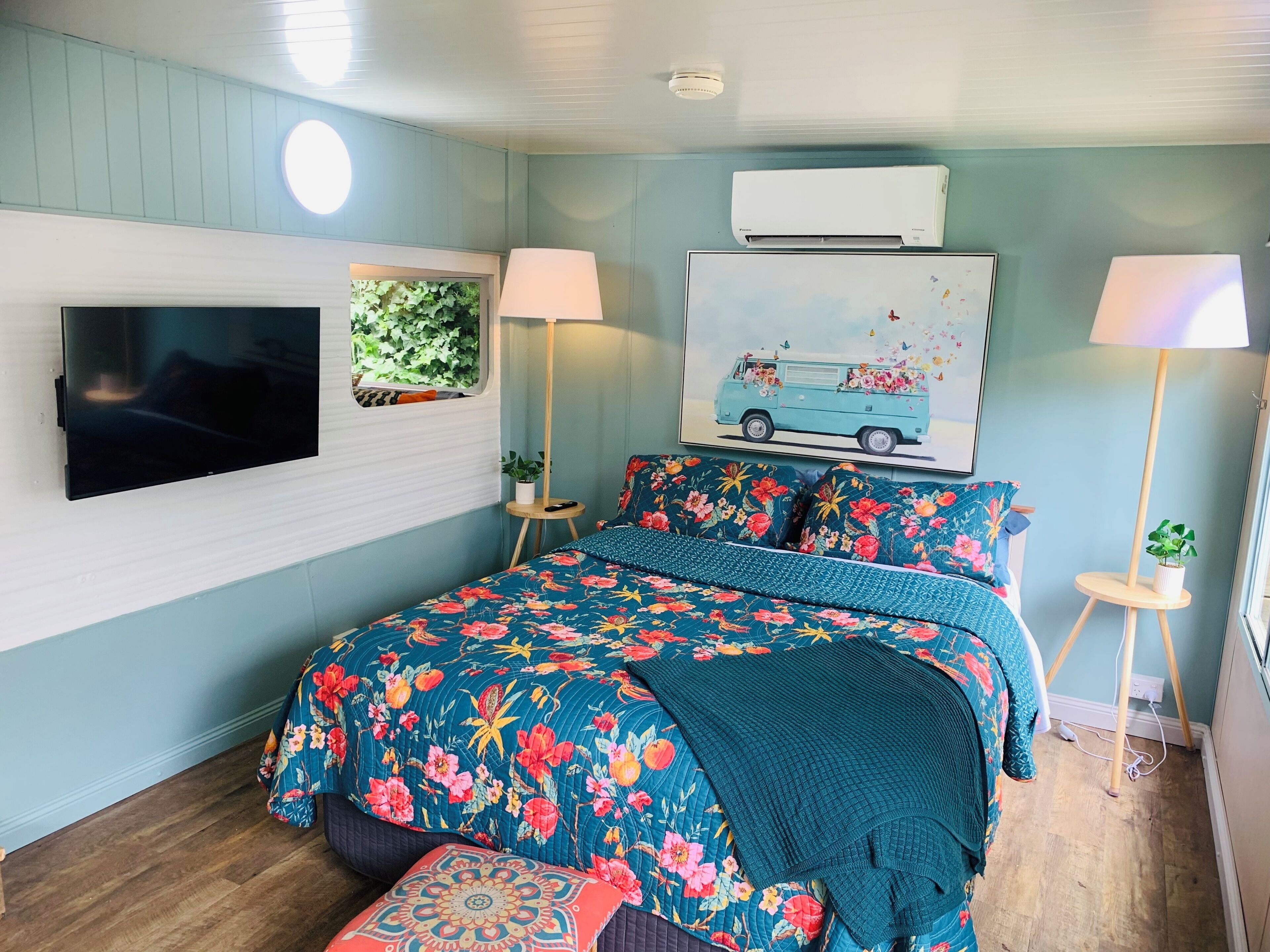 Quarto com cama, duas mesas de cabeceira, televisão, ar condicionado e janela com vista para o verde.