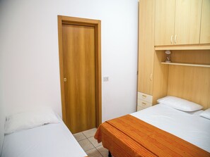 Quarto pequeno e limpo, com duas camas, um guarda-roupa de madeira e uma pequena mesa.