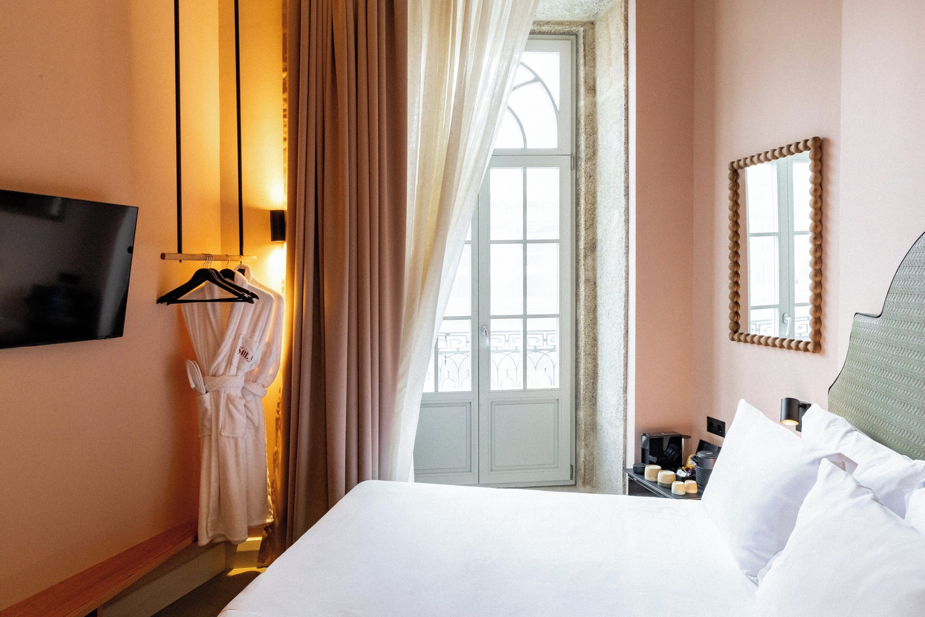 Quarto de hotel com cama, televisão, roupão, espelho e janela com cortinas.