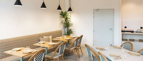 Uma sala de jantar com assentos tipo camarote e mesas, iluminação pendente, uma planta em vaso e uma porta que dá para a cozinha.
