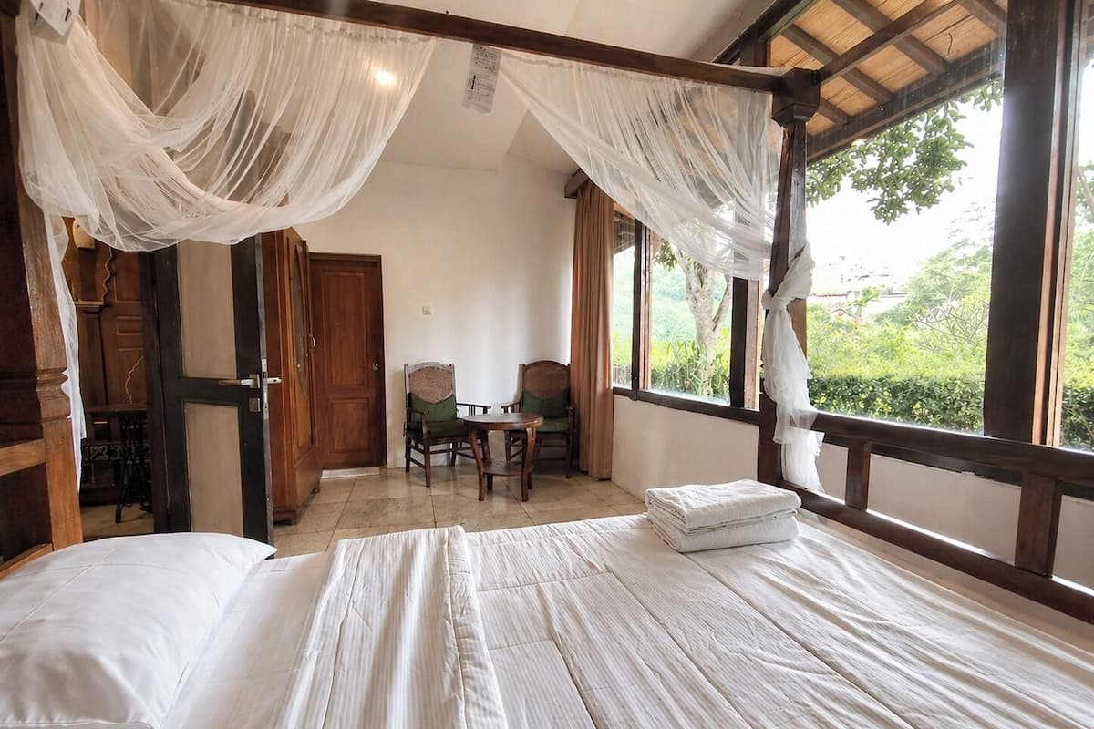 Quarto com cama, mesa, duas cadeiras e janela com vista para o verde.