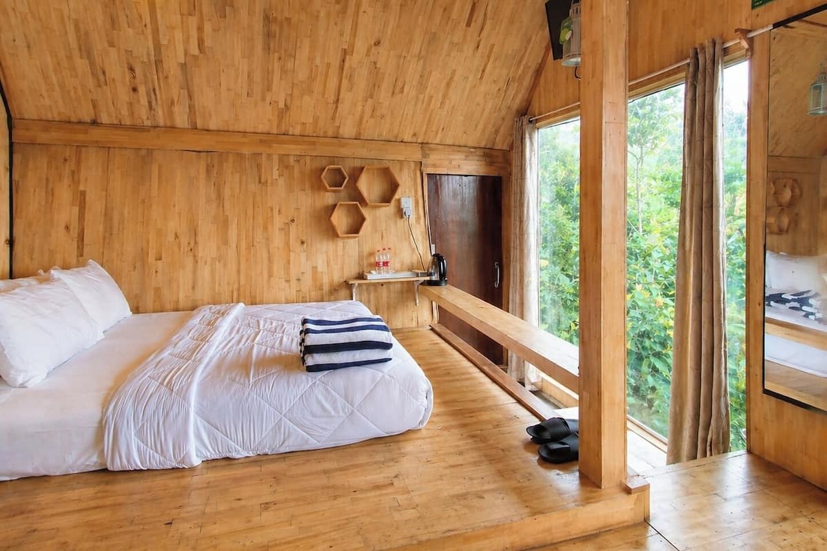 Quarto de cabana de madeira com uma cama, uma pequena mesa, uma lâmpada pendente e uma janela com cortinas.