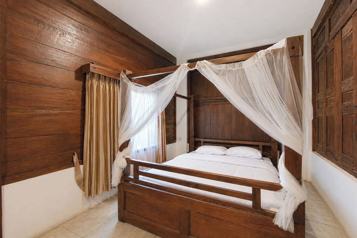Uma cama de madeira com dossel, roupa de cama branca e cortinas leves, apoiada em uma parede e piso de madeira.