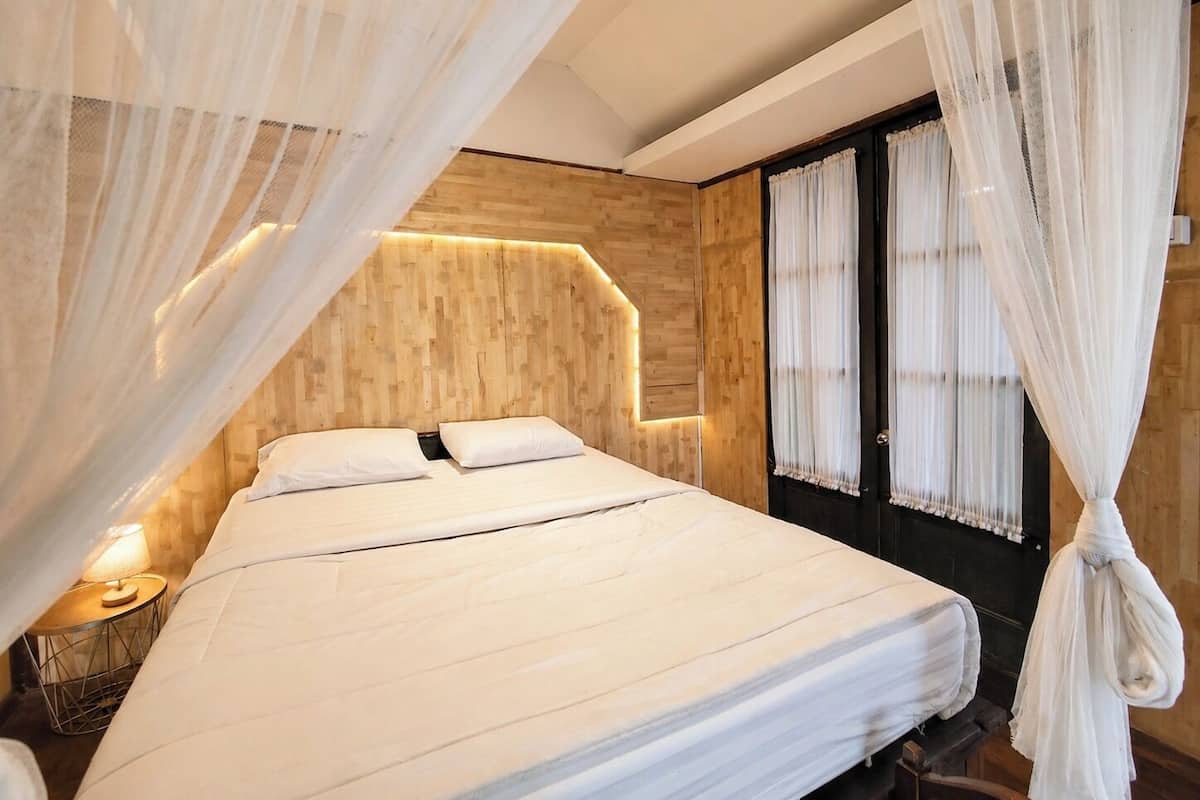 Quarto com cama, mesa de cabeceira, luminária e janela com cortinas.