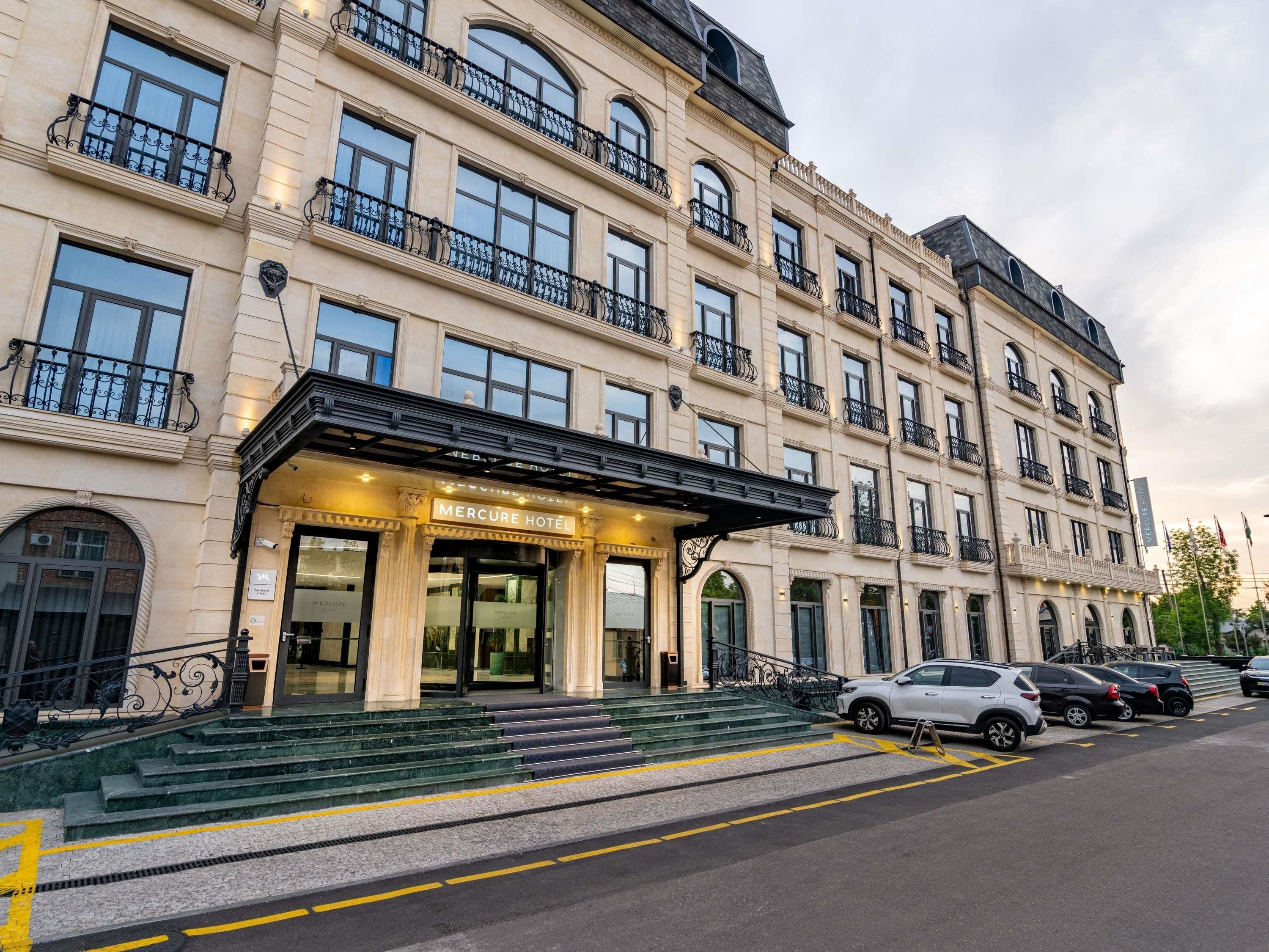 Um hotel de vários andares com fachada clássica, uma entrada imponente e um estacionamento com vários carros.