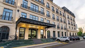 Um hotel de vários andares com fachada clássica, uma entrada imponente e um estacionamento com vários carros.