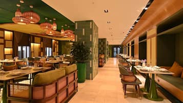 Restaurante com pé-direito alto, luminárias de vime pendentes e uma planta verde central. Mesas com toalhas brancas e cadeiras verdes.