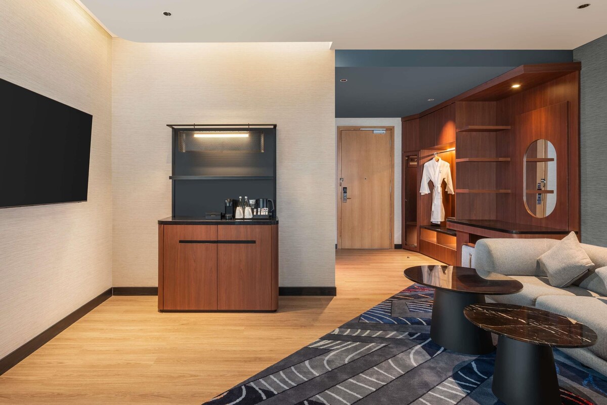 Quarto de hotel moderno com um armário de madeira, TV de tela plana, área de estar com sofá e mesa de centro, e um roupão pendurado em um guarda-roupa de madeira.