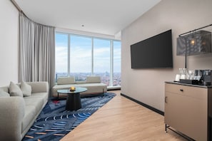 Quarto de hotel moderno com sofá, mesa de centro, TV na parede e vista para a cidade.
