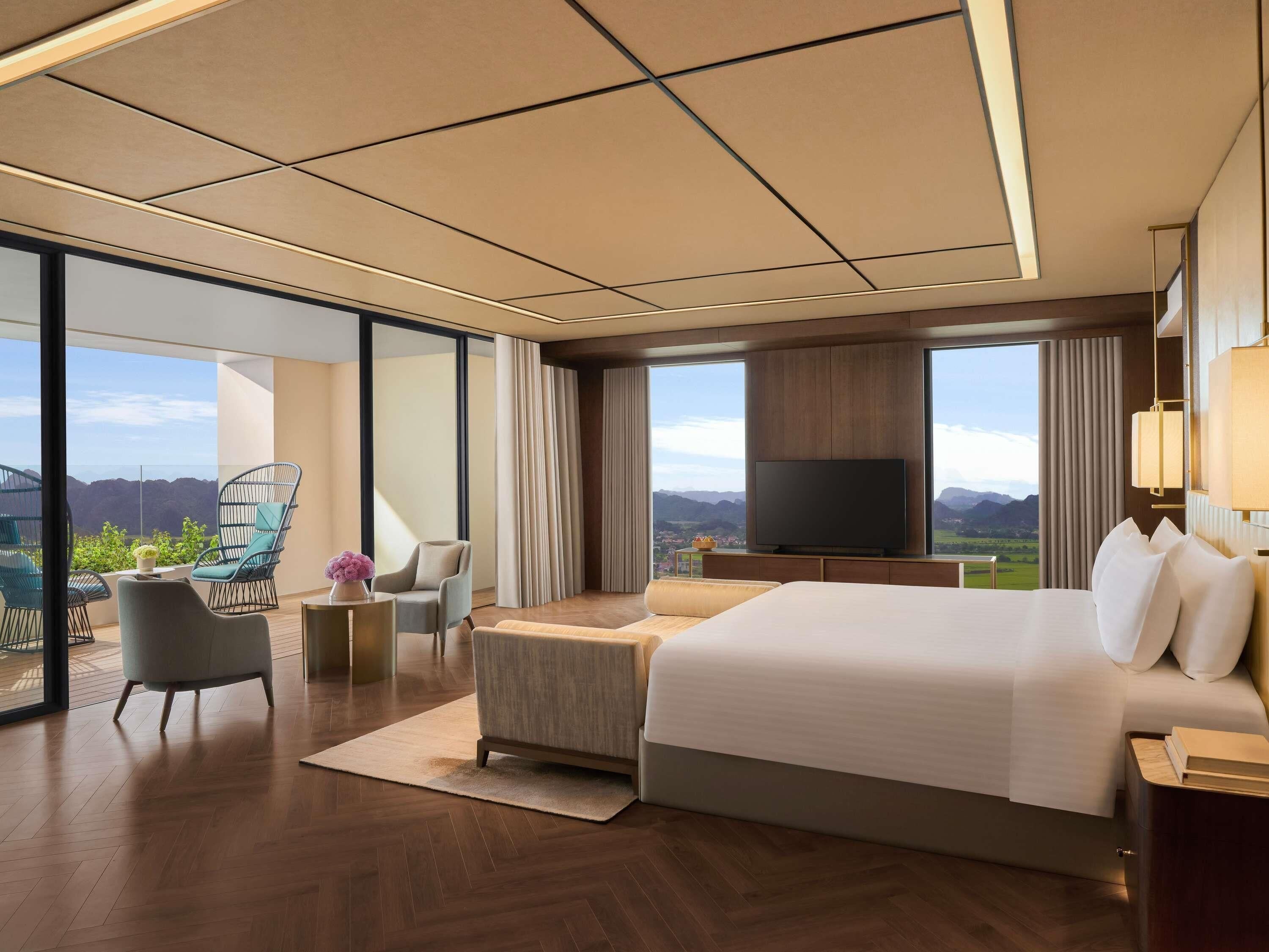 Quarto de hotel moderno com uma cama grande, televisão, área de estar com cadeiras, uma mesinha e vista para as montanhas.