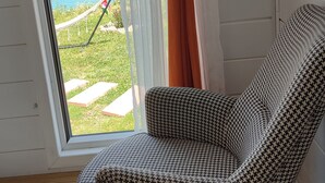 Quarto com uma cadeira de padrão xadrez, uma janela grande com cortinas brancas e vista para a paisagem costeira.