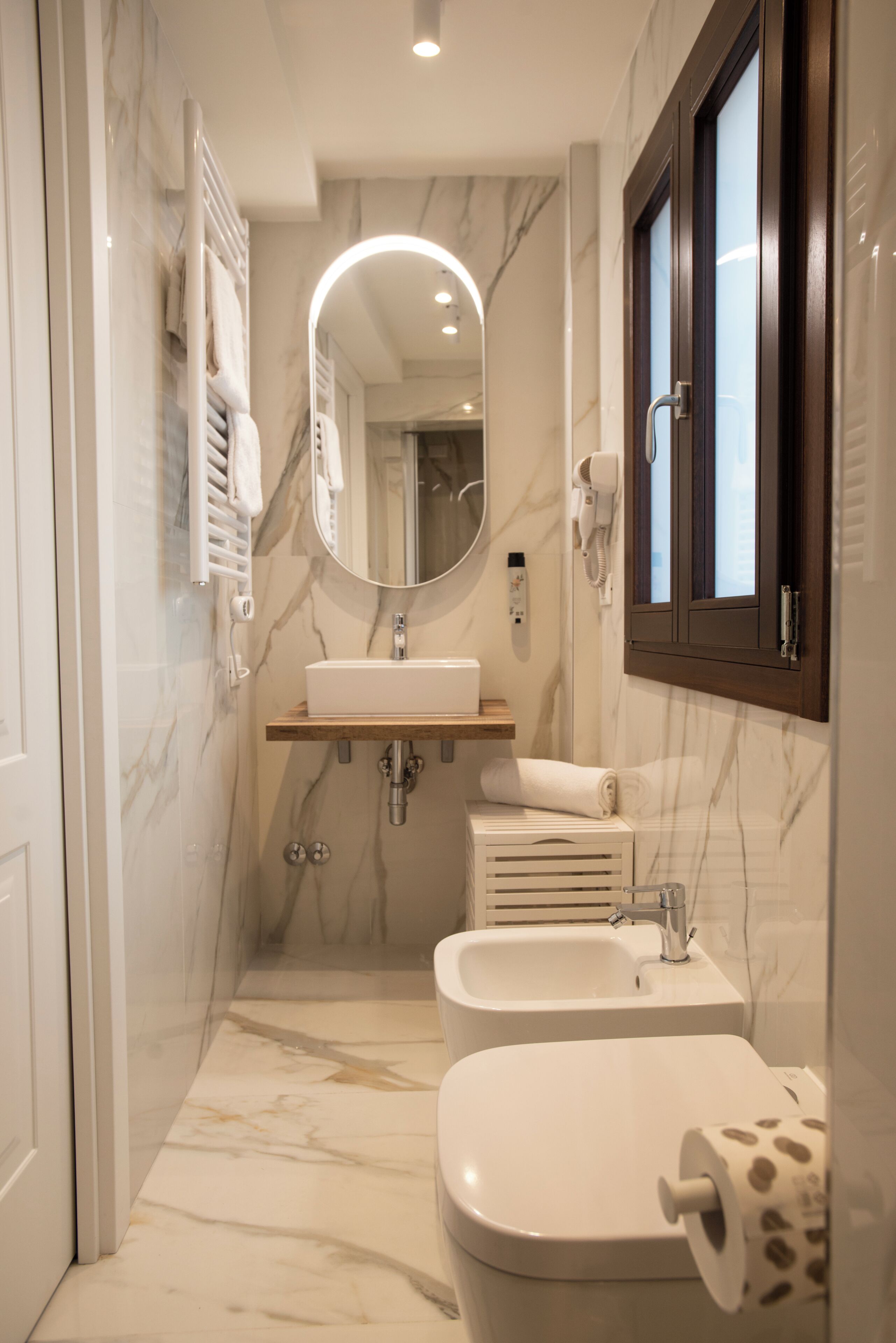 Uma casa de banho moderna com soalho de mármore, uma sanita branca, um bidé, uma casa de wc, um porta-toalhas e um espelho com luz em cima.