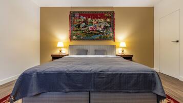 Quarto de hotel com uma cama grande, duas luminárias de cabeceira, tapeçaria na parede e um tapete vermelho.