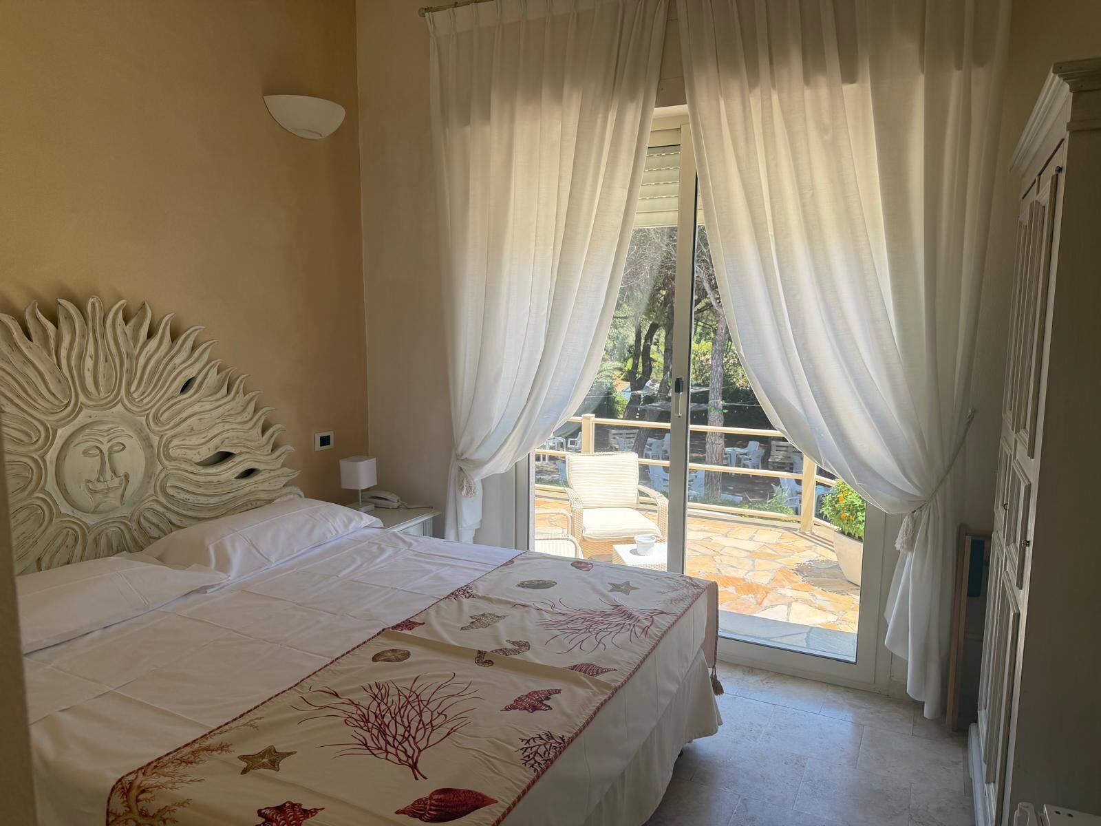 Quarto com cama grande, cabeceira em formato de sol, varanda com mobiliário exterior e cortinas leves.