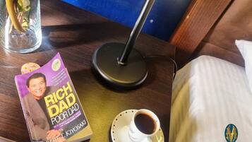 Quarto de hotel com uma mesa de cabeceira, um livro intitulado 'Rich Dad Poor Dad', uma chávena de café e um candeeiro.