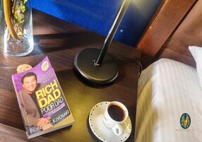 Quarto de hotel com uma mesa de cabeceira, um livro intitulado 'Rich Dad Poor Dad', uma chávena de café e um candeeiro.