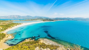 Paisagem costeira com mar azul cristalino, praia arenosa e montanhas ao longe.