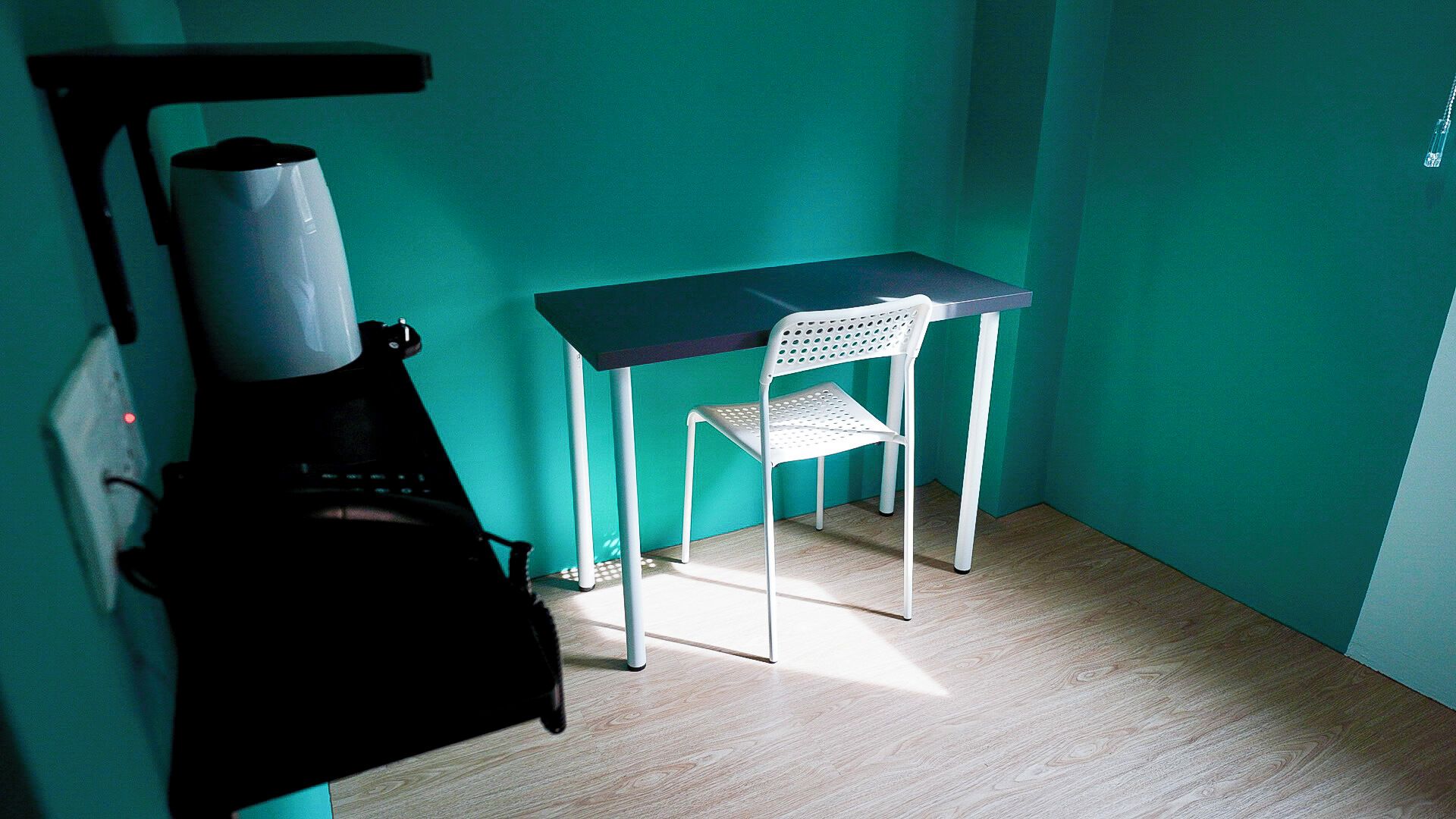 Quarto pequeno com paredes verde-água, piso de madeira, uma cadeira branca e um objeto preto à esquerda.
