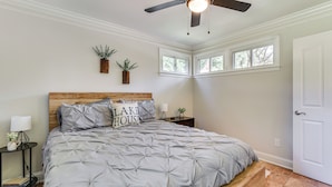 Quarto com cama, mesa de cabeceira, abajur, ventilador de teto e porta.