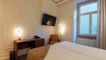 Quarto de hotel com cama, escrivaninha, cadeira, televisão na parede e janela com cortinas.