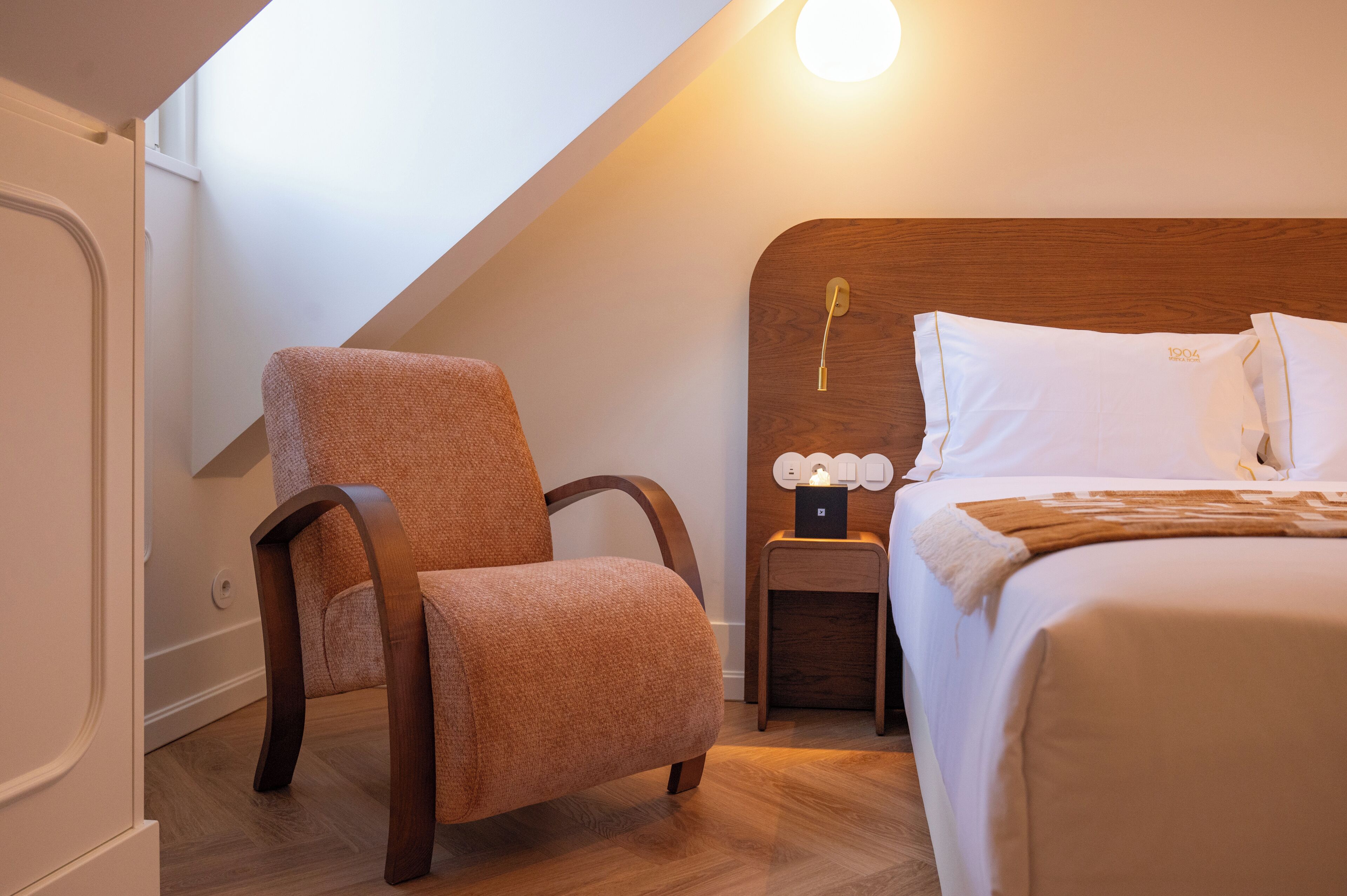Quarto de hotel com cabeceira de madeira, uma cama de casal, mesa de cabeceira com abajur, uma cadeira e janela com vista para o céu.