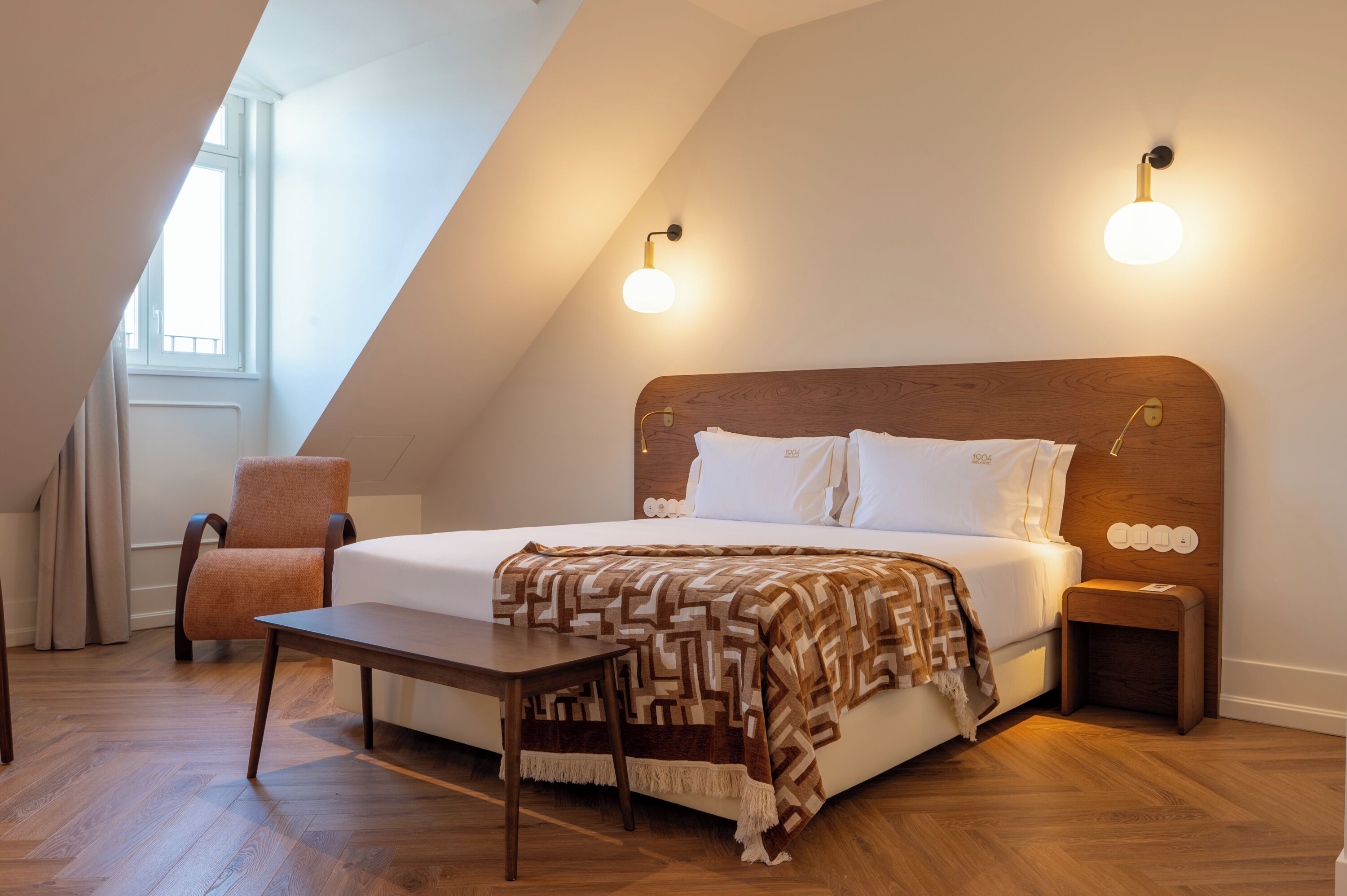 Quarto com cabeceira de madeira, uma cama grande, mesa de cabeceira, criadeira e uma cadeira.