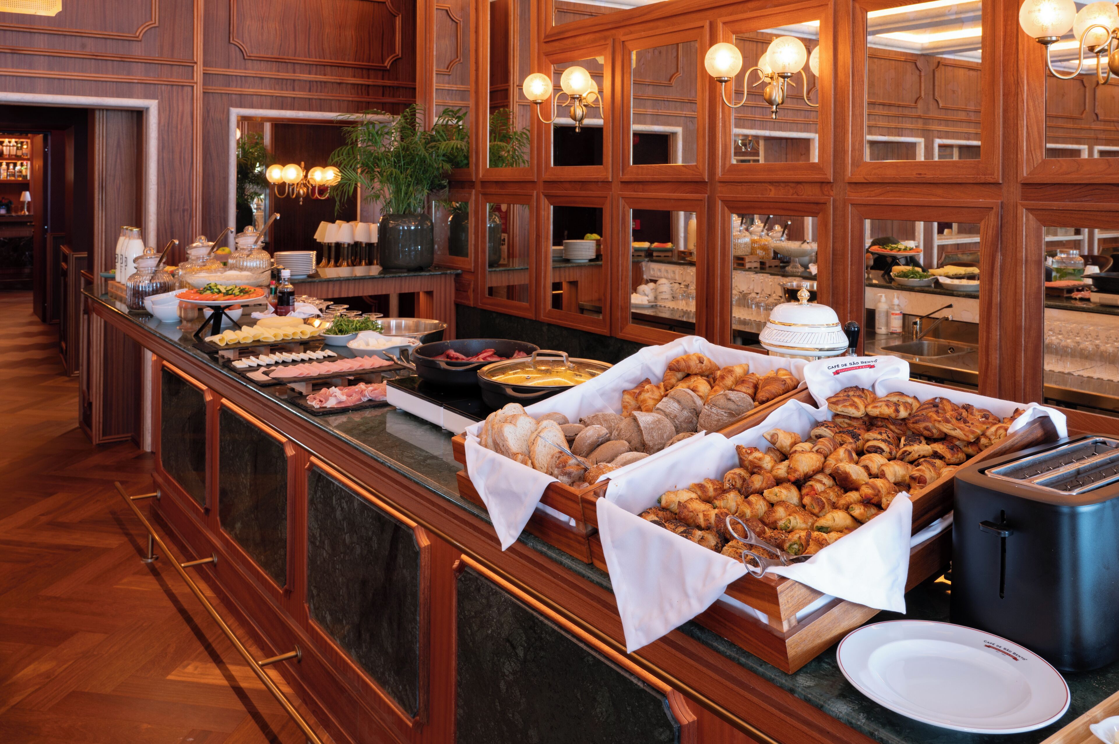 Buffet com diversos alimentos, incluindo doces e carnes, em um ambiente bem iluminado com decoração em madeira e bancada de mármore.