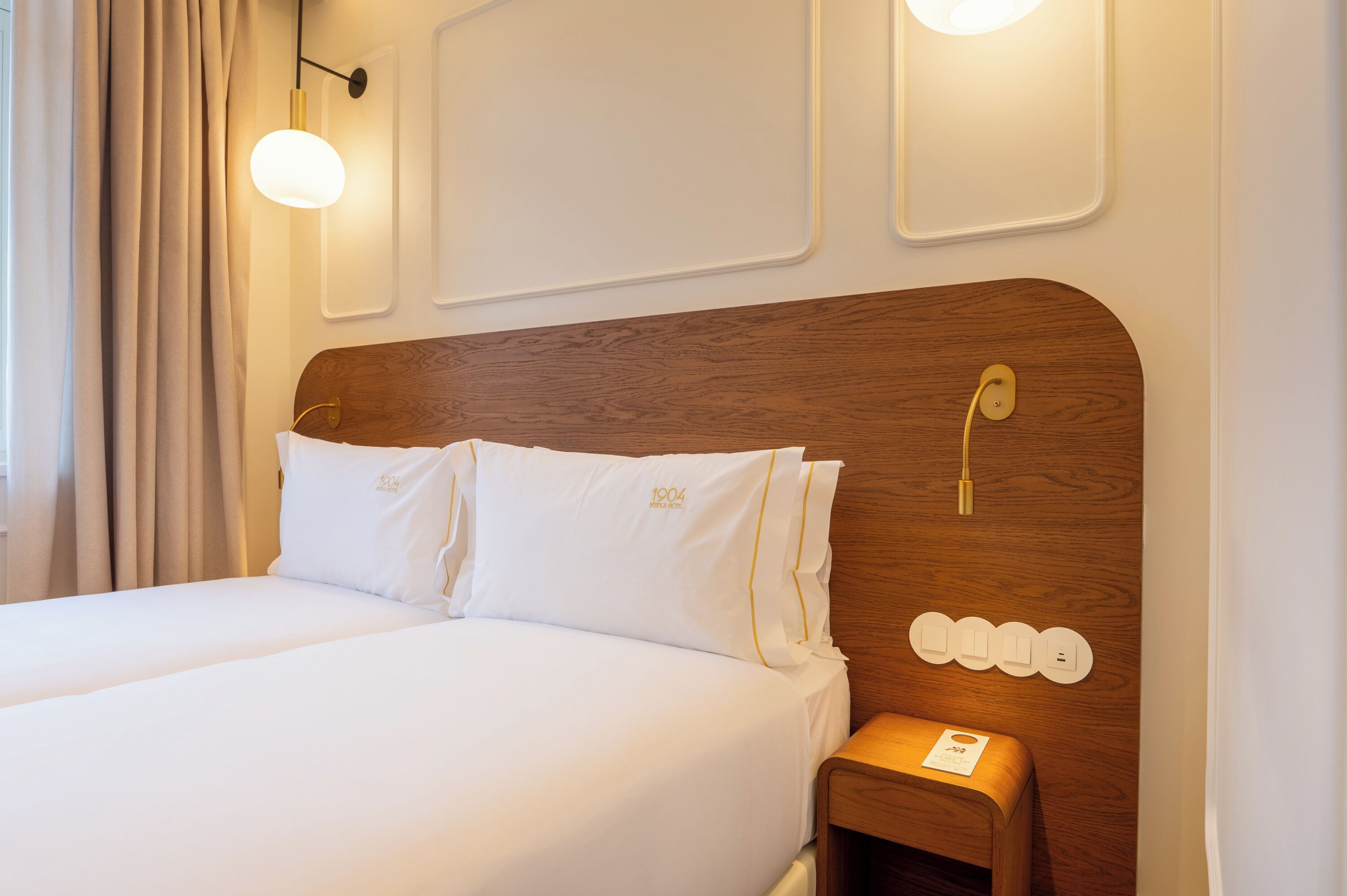 Quarto de hotel com cabeceira de madeira, duas almofadas brancas, mesa de cabeceira e luminárias fixadas na parede.
