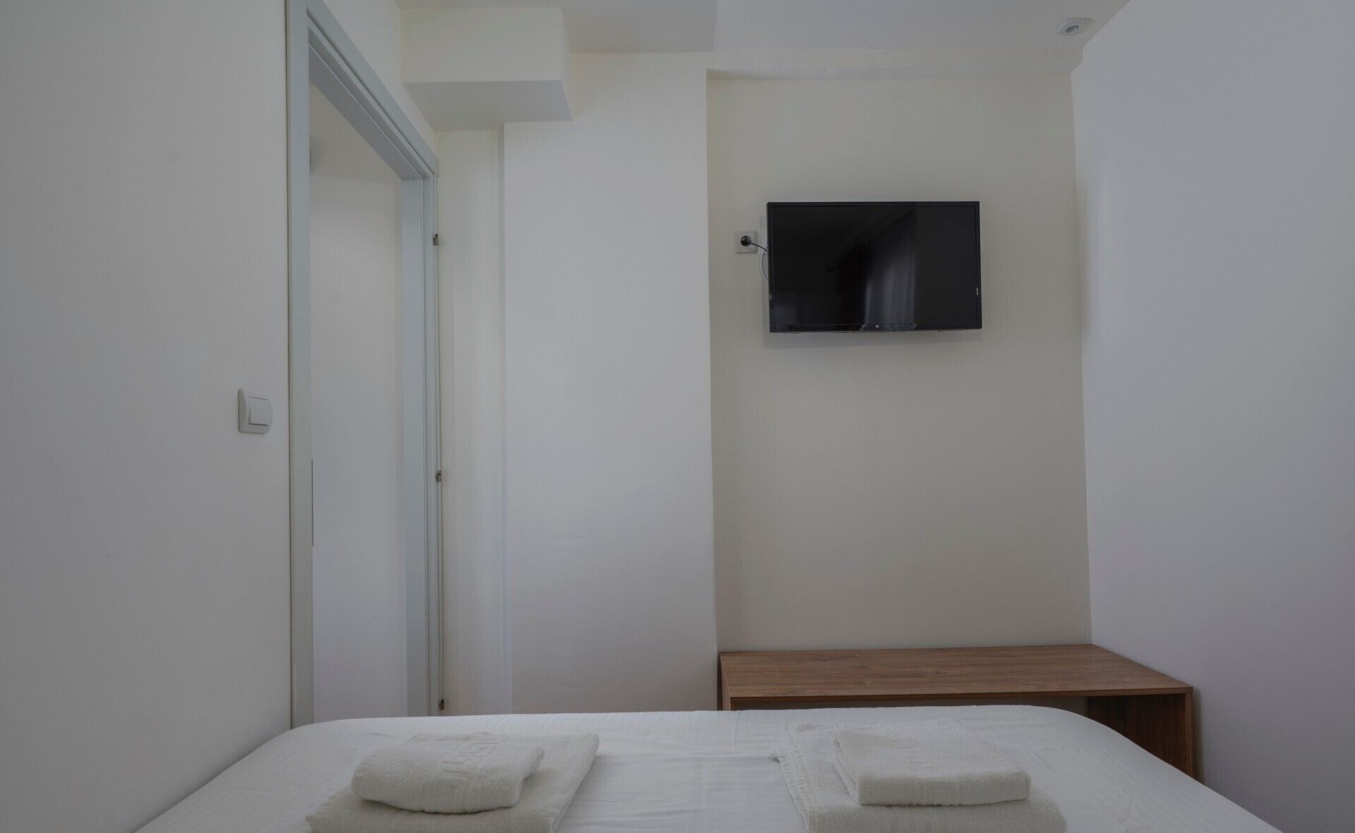 Um quarto pequeno e moderno com uma cama, mesa de cabeceira, TV na parede e uma porta.