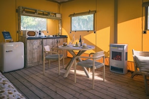 Quarto aconchegante com piso de madeira, uma pequena cozinha compacta com micro-ondas, geladeira e uma mesa com cadeiras. Há duas janelas que proporcionam iluminação natural, e é visível uma lareira a lenha.