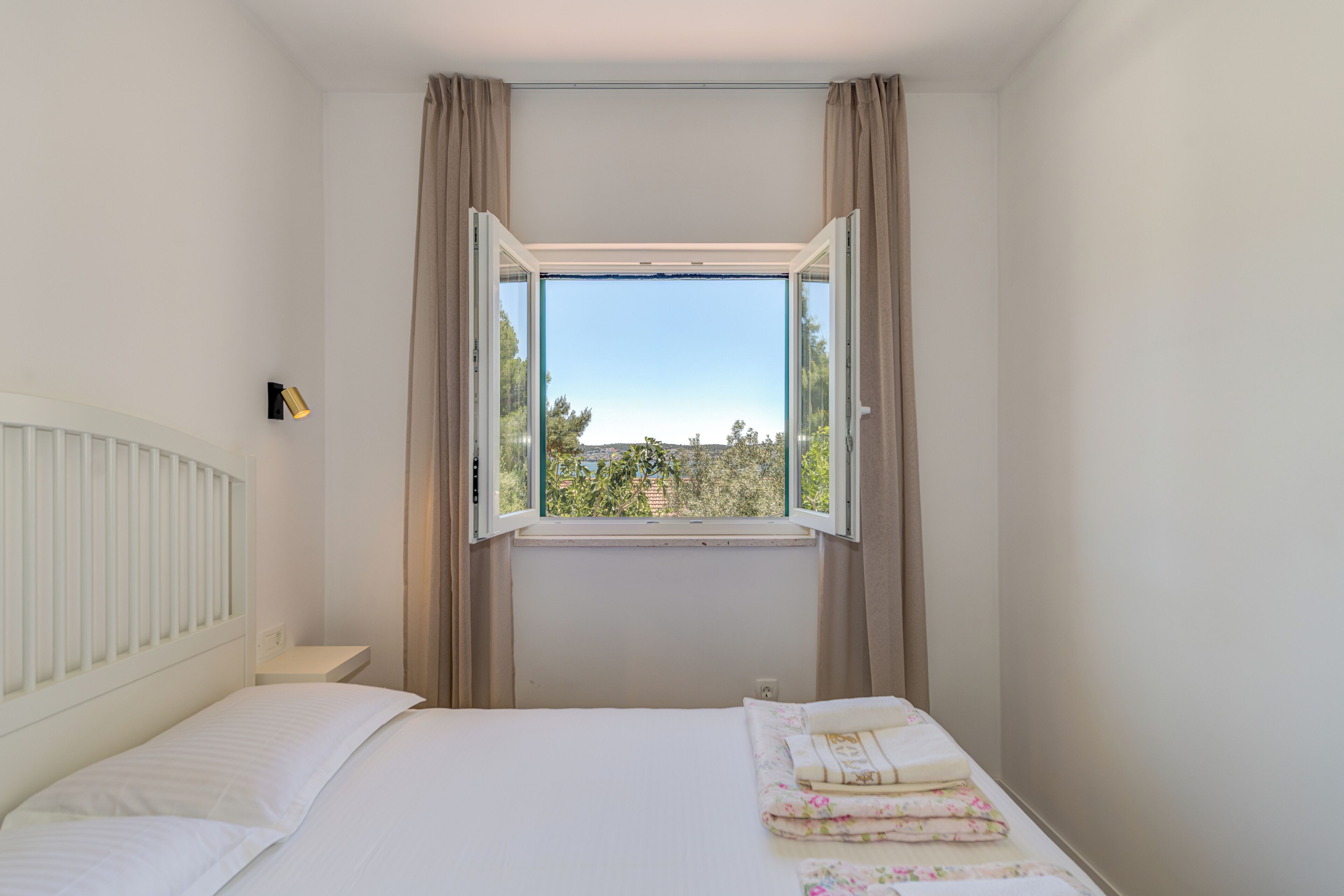 Quarto com cama, janela com cortinas, luminária de cabeceira e vista para o verde lá fora.