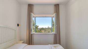Quarto com cama, janela com cortinas, luminária de cabeceira e vista para o verde lá fora.