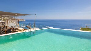 Céu azul e límpido, piscina com vista para o mar e área de estar com mesas e cadeiras.