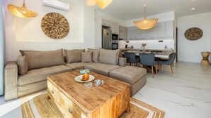 Sala de estar moderna com sofá seccional, mesa de centro de madeira e área de refeições, com cozinha ao fundo.