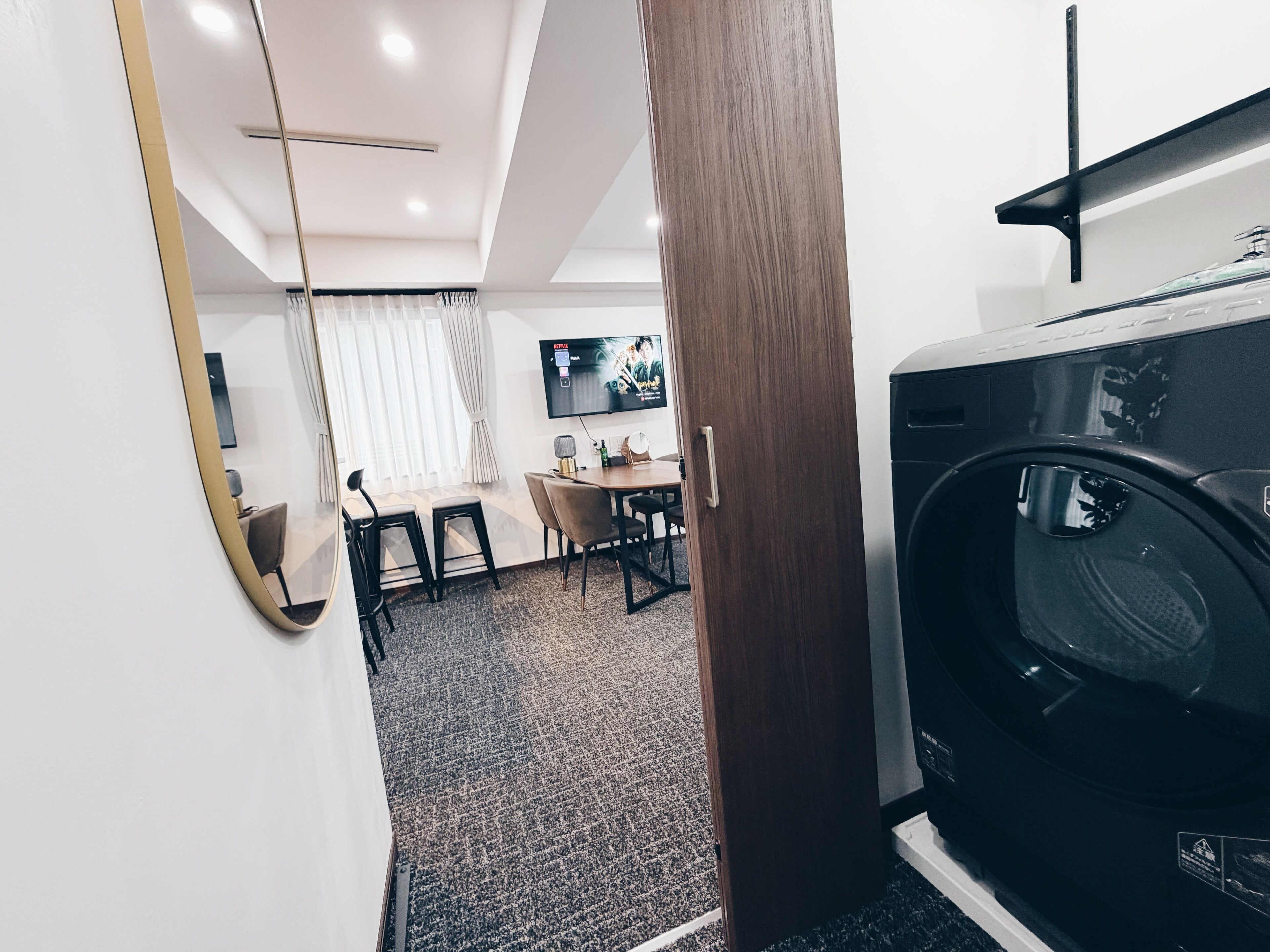 Quarto de hotel com máquina de lavar roupa, área de refeições com mesa e cadeiras, e televisão na parede.