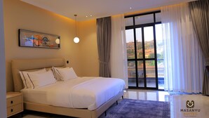 Quarto totalmente mobiliado com cama grande, mesas de cabeceira, luminária no teto, janela com cortinas e vista para a cidade.