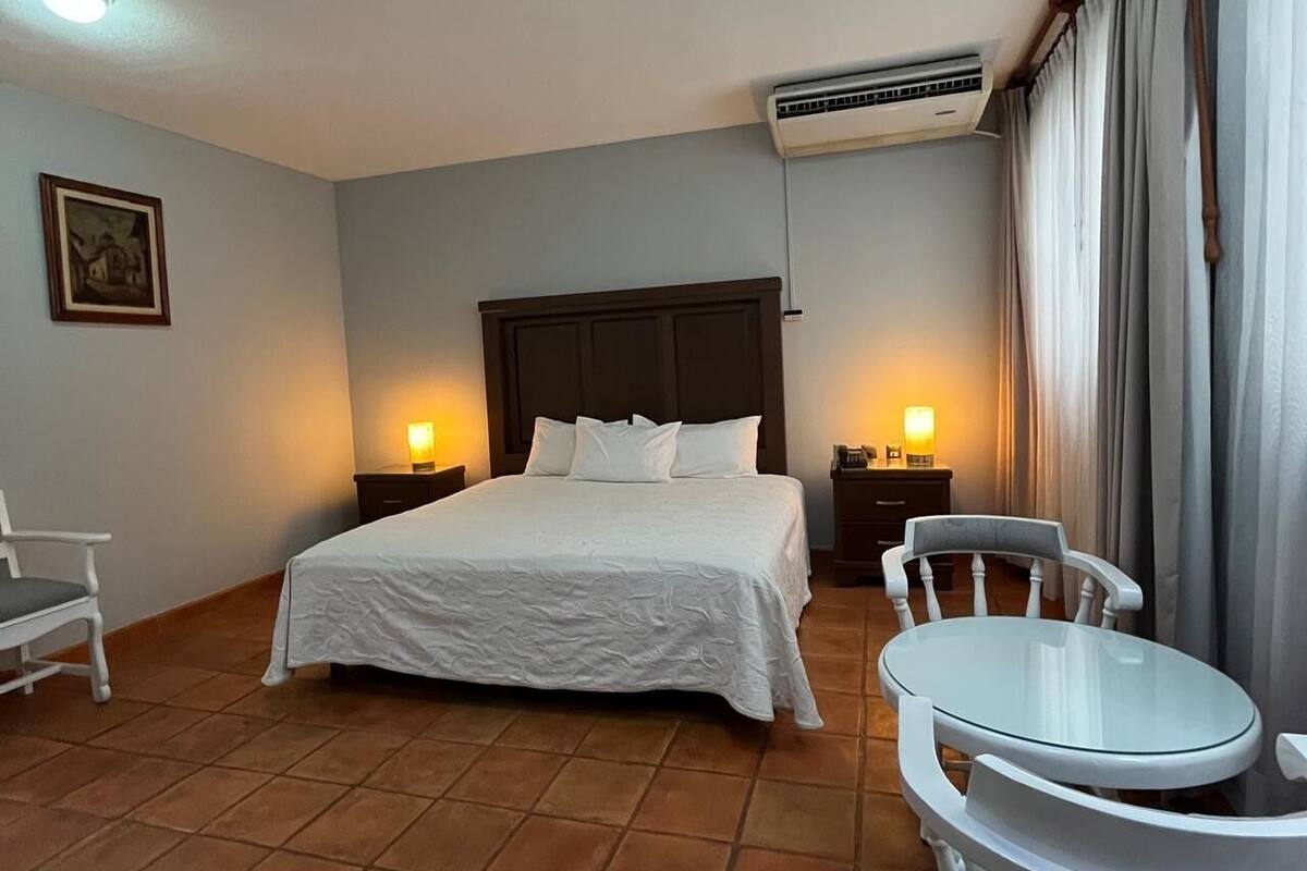 Quarto de hotel com uma cama grande, duas mesas de cabeceira com luminárias, uma pequena mesa redonda com cadeiras e um aparelho de ar condicionado na parede.