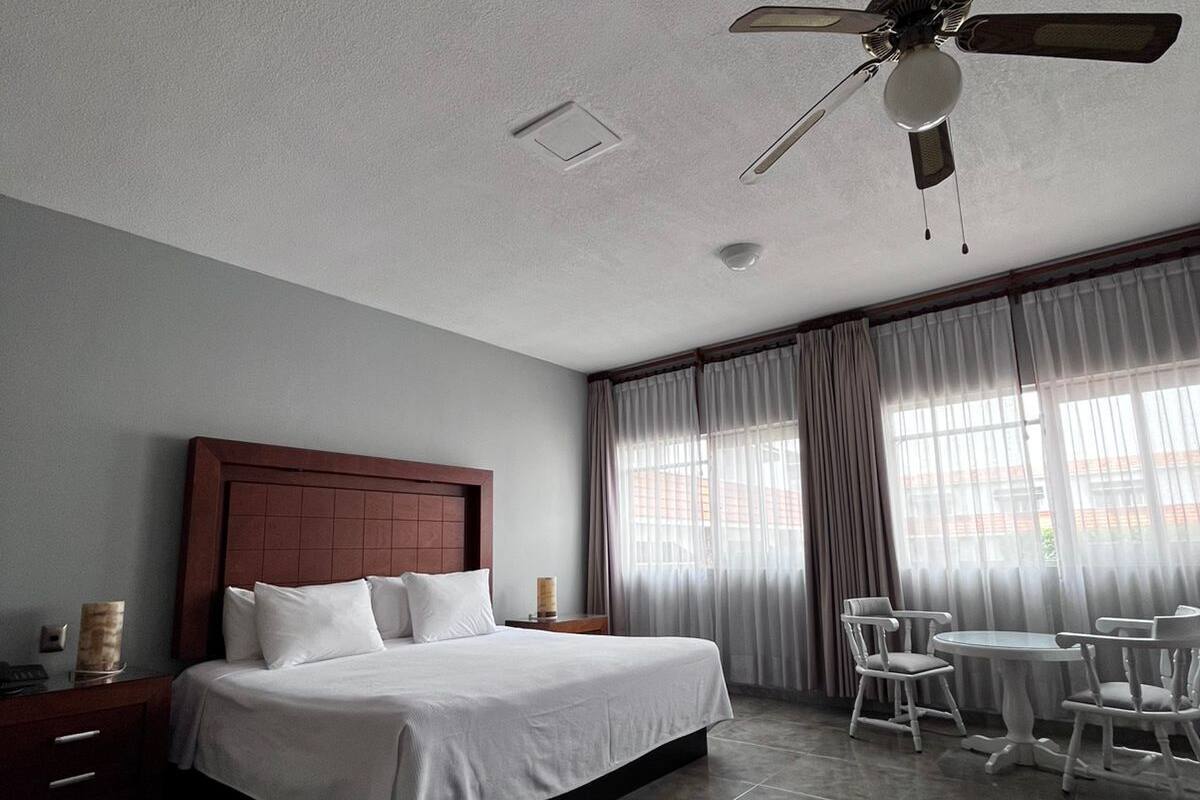 Quarto de hotel com uma cama grande, um ventilador de teto, uma mesinha com cadeiras e vista para o exterior através de amplas janelas.