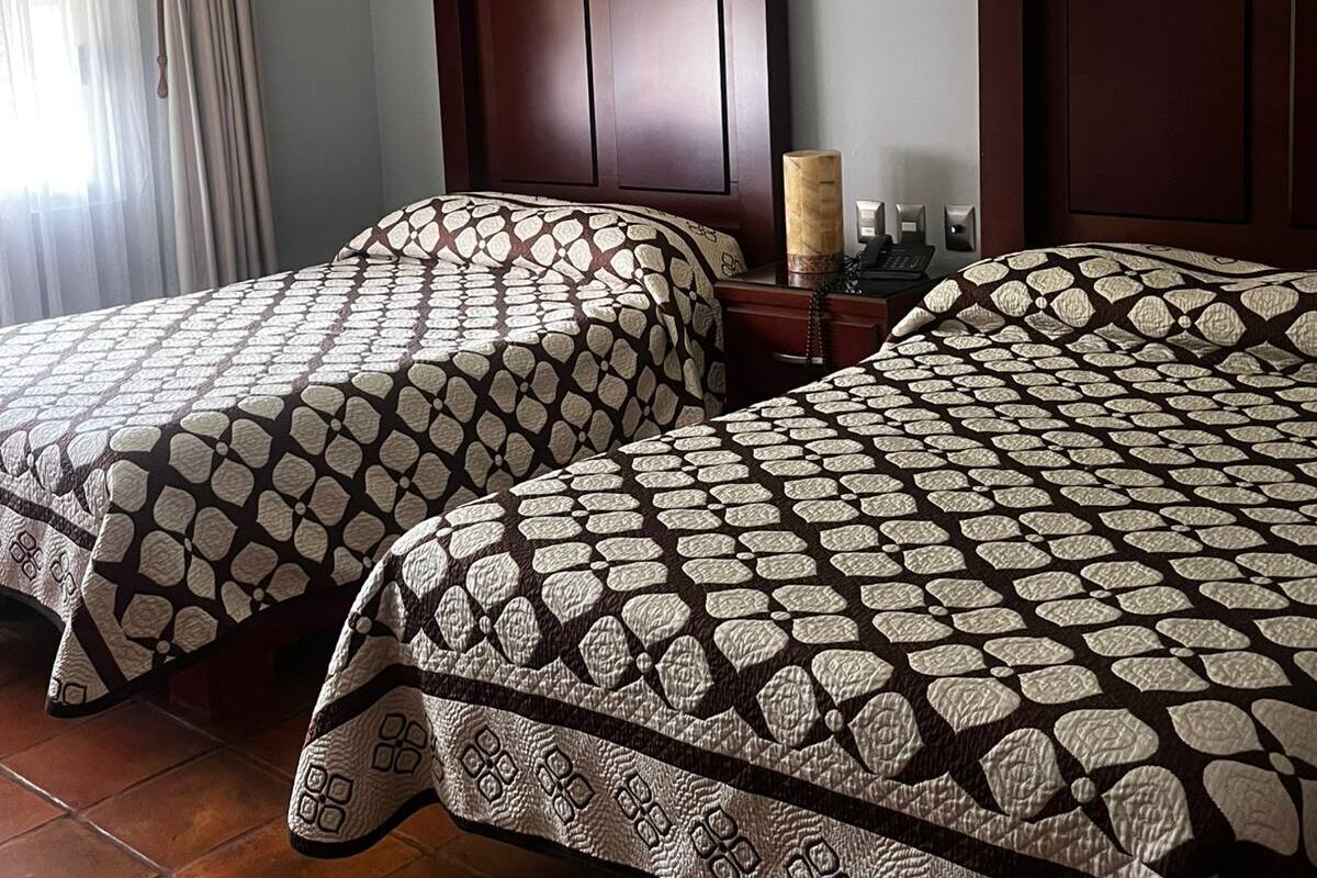 Quarto de hotel com duas camas, um ventilador de teto, cabeceira de madeira e mesa de cabeceira com abajur.