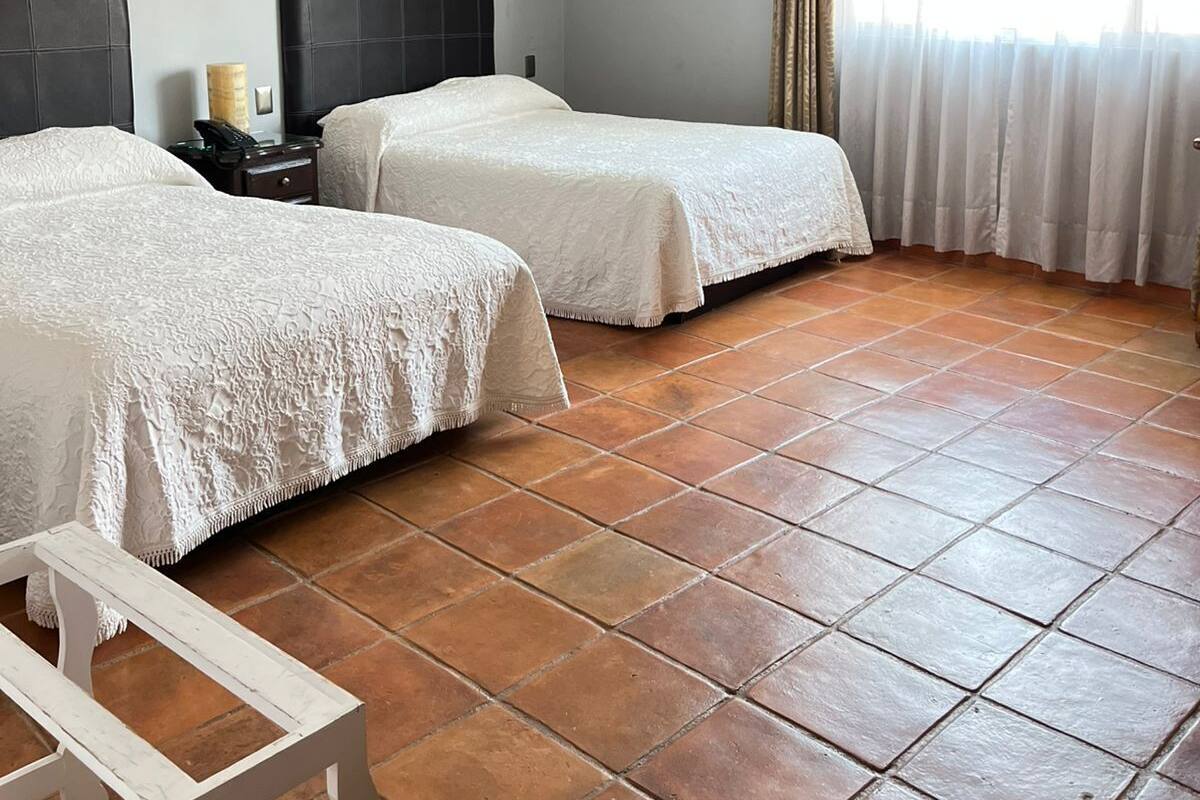 Quarto de hotel com duas camas, um ventilador de teto, um ar condicionado, uma mesa de cabeceira com abajur e uma janela com cortinas.